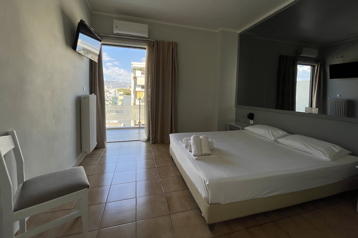 Quarto de hotel com cama, uma cadeira, TV fixada na parede e janela com cortinas.