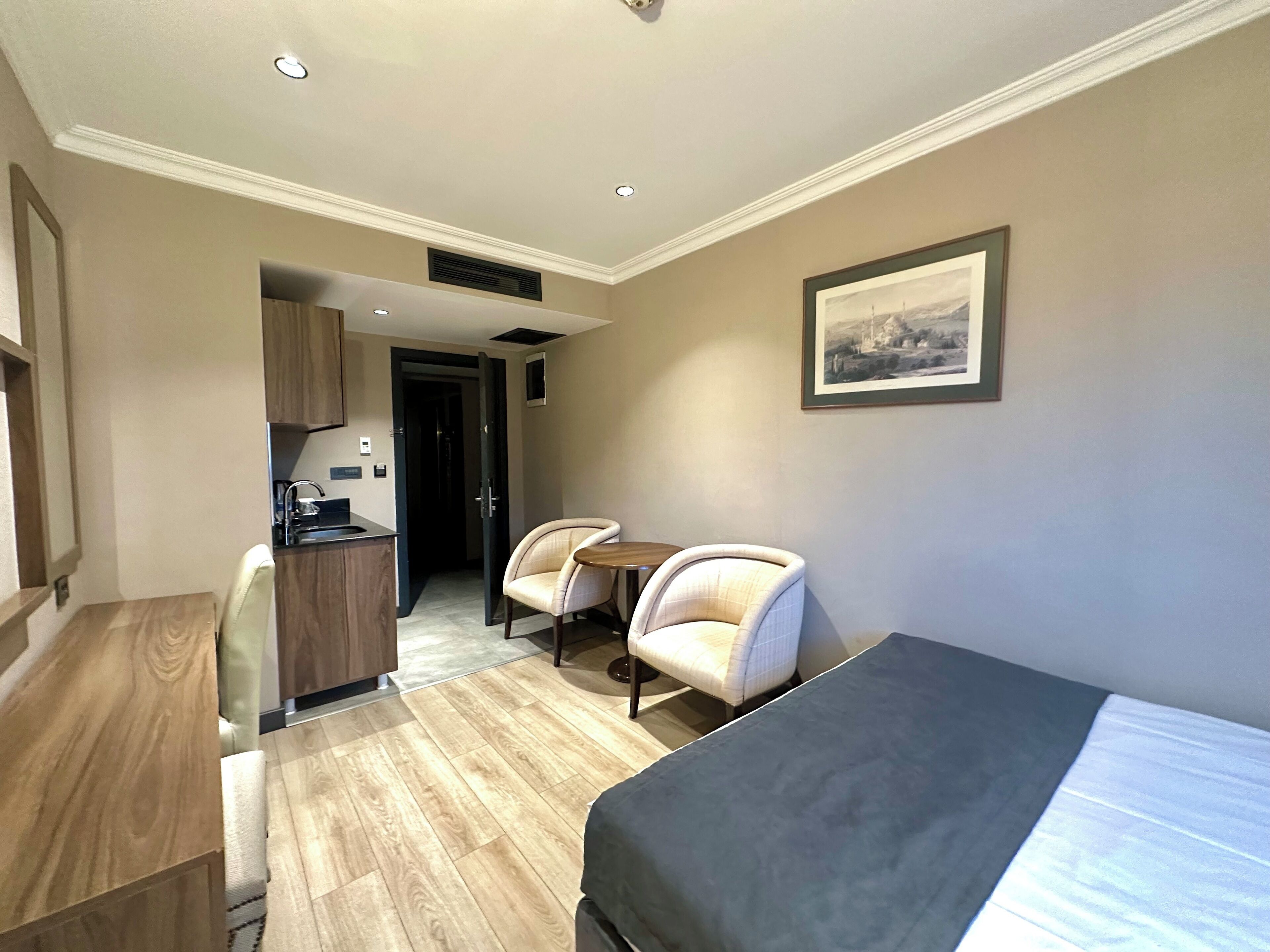 Quarto de hotel com cama, escrivaninha, cadeira, mesa pequena, cozinha compacta e porta que dá para outro cômodo.