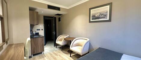 Quarto de hotel com cama, escrivaninha, cadeira, mesa pequena, cozinha compacta e porta que dá para outro cômodo.