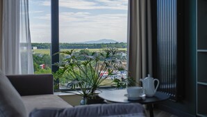 Quarto com uma janela grande, cama, mesa com chaleira e xícara, e um vaso com planta.