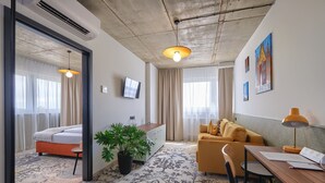 Quarto de hotel com cama, sofá, escrivaninha com luminária, planta em vaso e televisão.