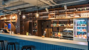 Uma área de bar com balcão, banquetas, prateleiras com garrafas, copos e um refrigerador.