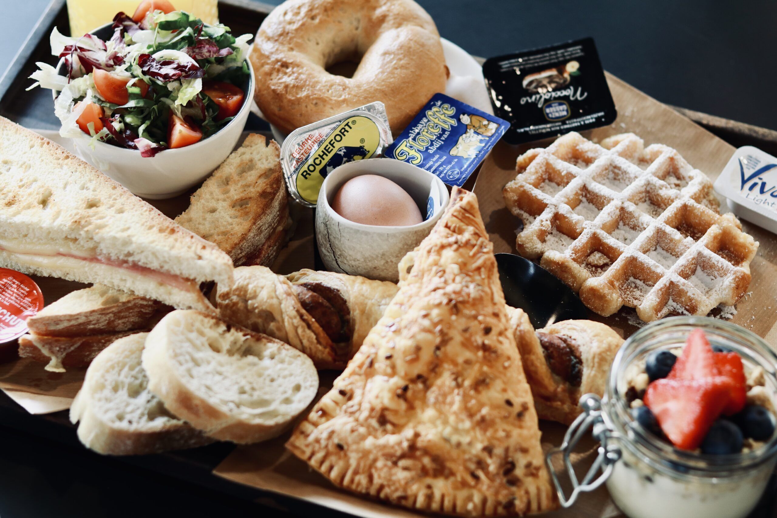 Uma bandeja com diversos itens de café da manhã, incluindo bagels, sanduíches, um waffle e uma salada.