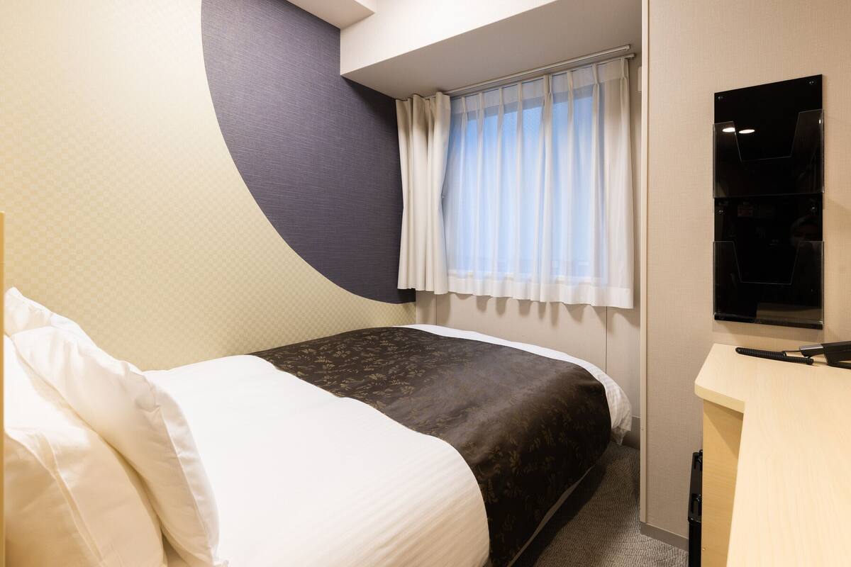 Quarto de hotel com cama, escrivaninha, televisão e janela com cortinas.