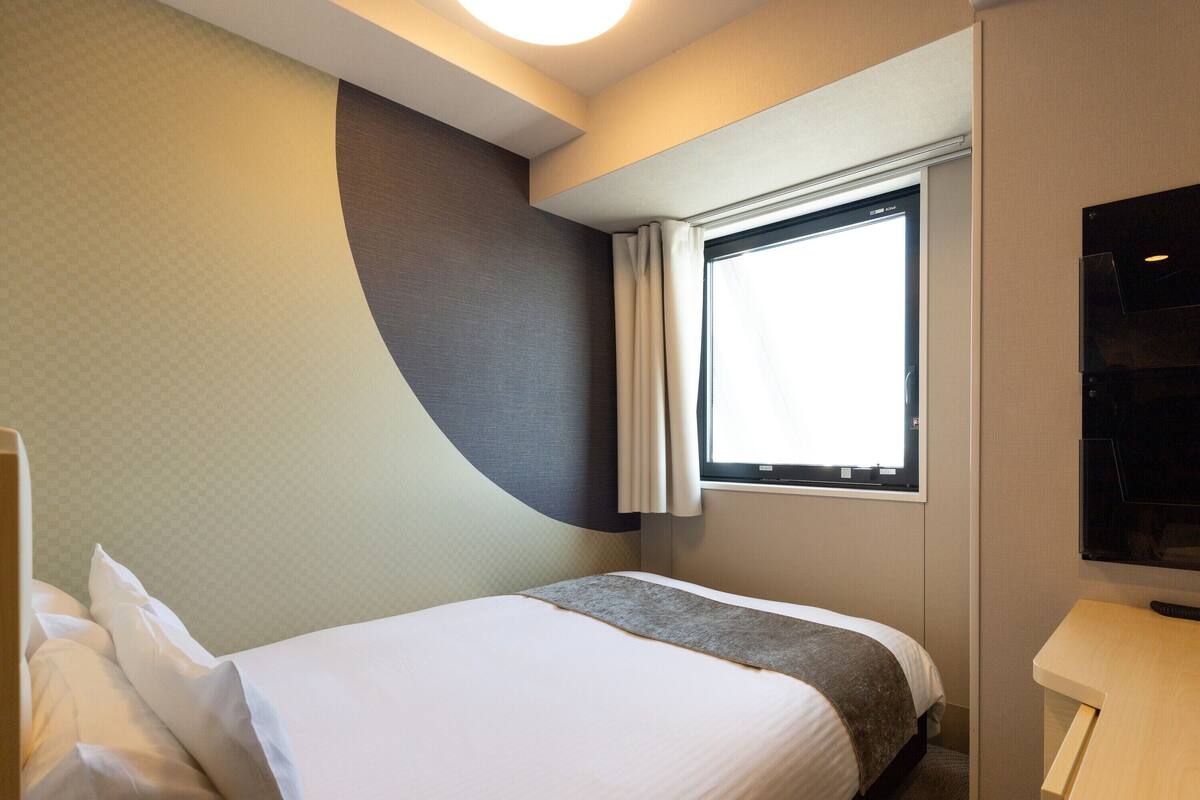 Quarto de hotel com cama, escrivaninha, janela com cortinas e televisão de tela plana na parede.