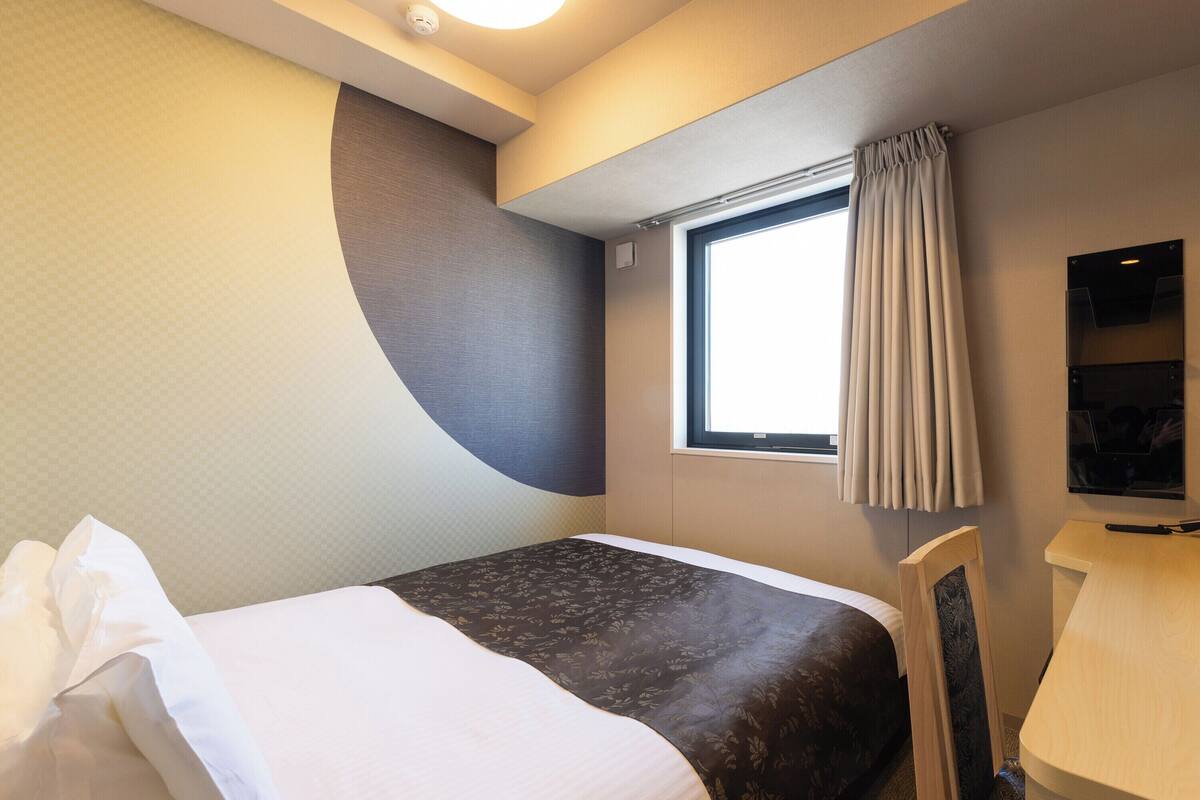 Quarto de hotel com uma cama, uma escrivaninha, uma janela com cortinas e uma luminária no teto.