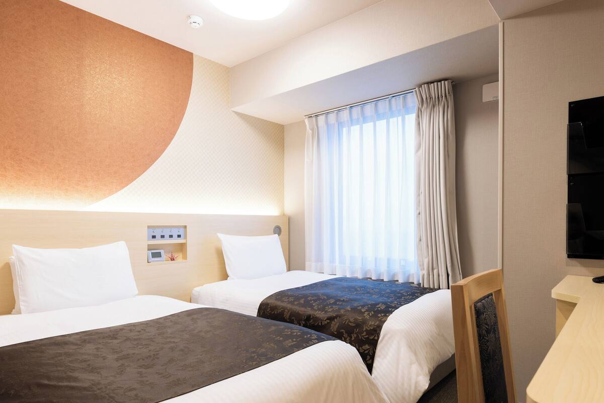 Quarto de hotel com duas camas, uma escrivaninha e uma janela grande com cortinas.
