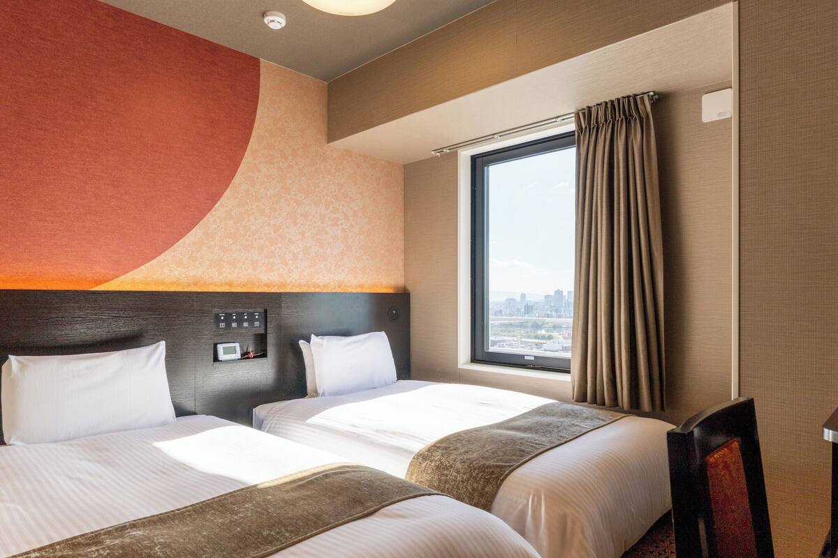 Quarto de hotel com duas camas, painel de controle na cabeceira, janela com cortinas e vista para a cidade.