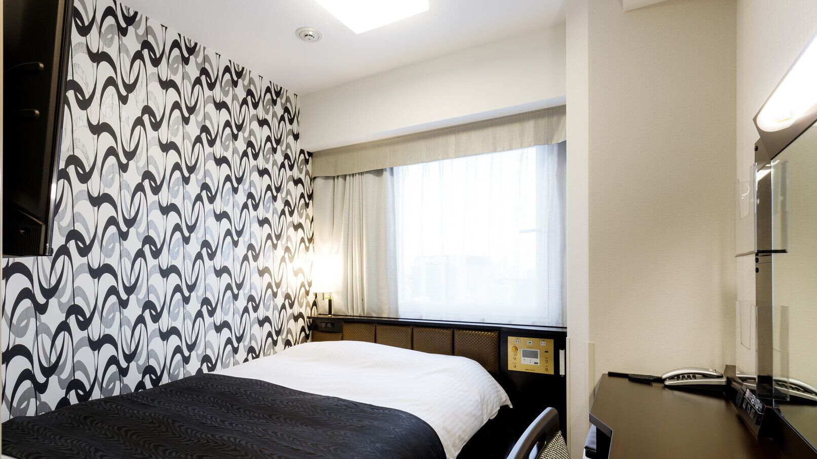 Quarto de hotel com uma cama, uma mesa com telefone, uma janela com cortinas e um papel de parede estampado.