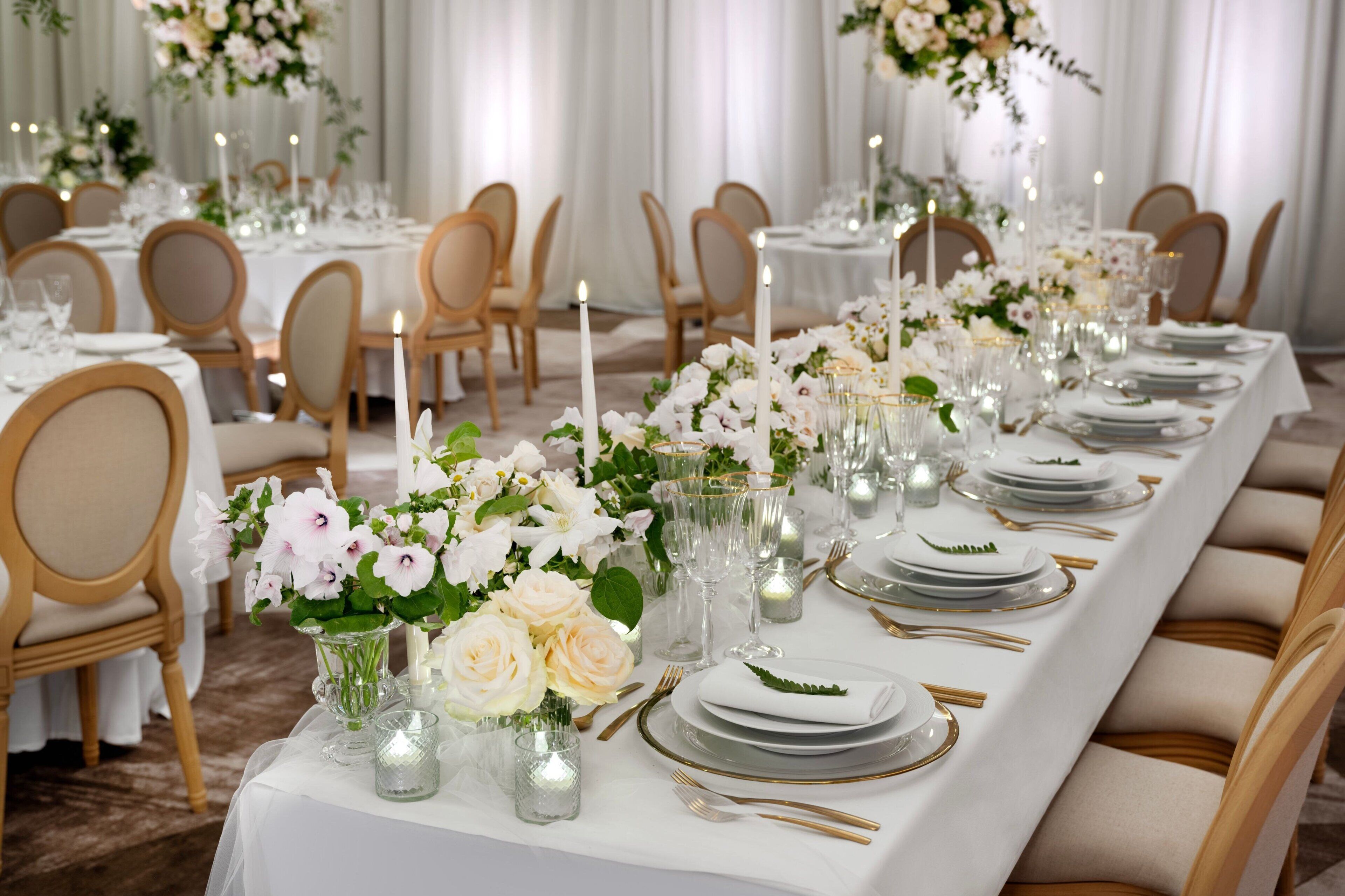 Uma sala de banquetes com longas mesas preparadas para um evento formal, com toalhas brancas, arranjos florais e iluminação de velas.