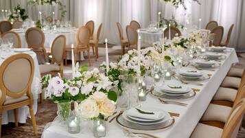 Uma sala de banquetes com longas mesas preparadas para um evento formal, com toalhas brancas, arranjos florais e iluminação de velas.
