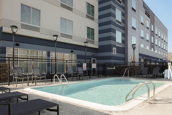 Área de piscina de hotel com espreguiçadeiras, piscina e um edifício com várias janelas e um letreiro.