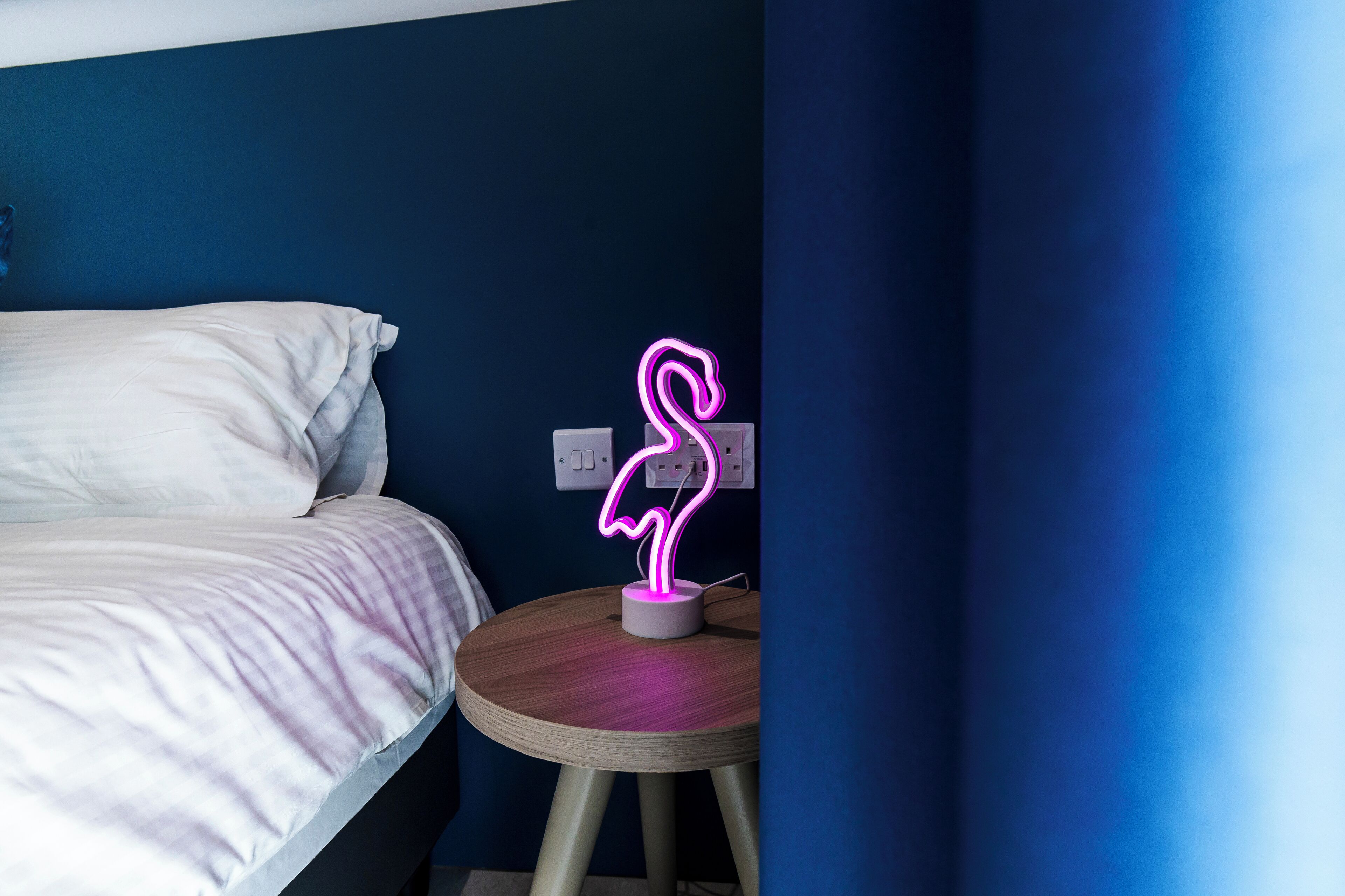 Quarto com cama, mesa de madeira redonda e um letreiro de néon em formato de flamingo.