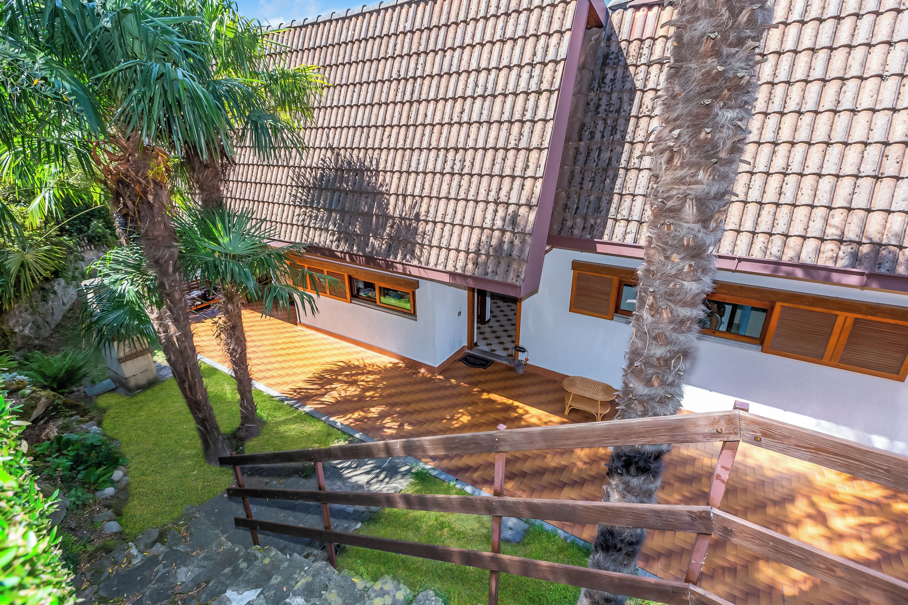 Um edifício de dois andares com telhado de telhas, escadaria de madeira e um jardim com palmeiras.