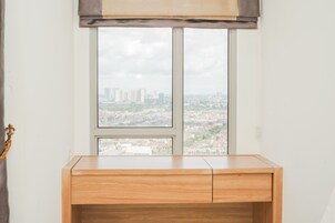 Quarto com escrivaninha de madeira e grande janela com vista para a paisagem urbana.