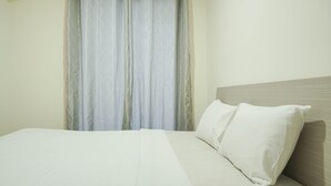 Uma cama bem arrumada com lençóis e almofadas brancos, cabeceira de madeira e uma janela com cortinas leves.