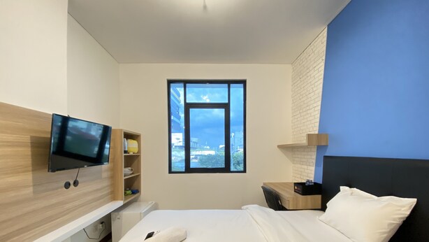 Quarto de hotel moderno com uma cama grande, uma televisão de tela plana na parede, uma janela com vista para a cidade e uma prateleira de madeira com objetos decorativos.