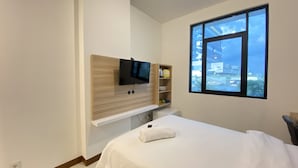 Quarto de hotel moderno com uma cama, televisão montada, janela com vista para a cidade e um pequeno estante com artigos de higiene pessoal.