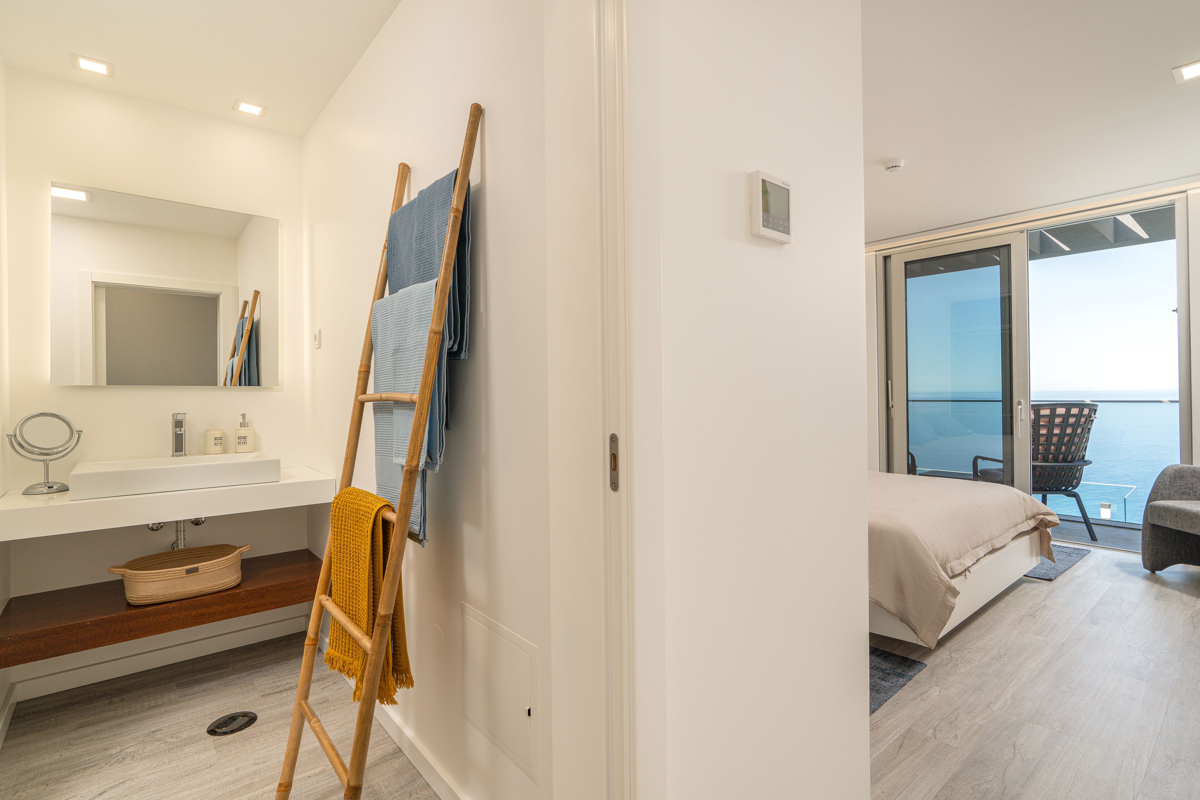 Quarto de hotel moderno com uma janela grande, cama, banheiro com espelho e pia, e uma escada de madeira.