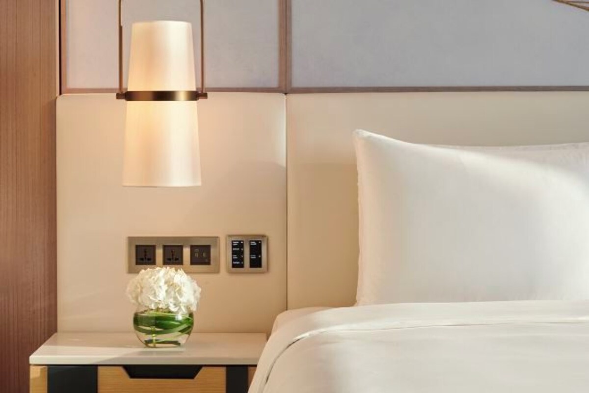 Quarto de hotel com cama, mesa de cabeceira com um vaso de flores, luminária na parede e painel de controle com botões.
