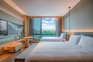 Quarto de hotel moderno com duas camas, uma janela ampla com vista para a cidade, área de refeições e televisão na parede.
