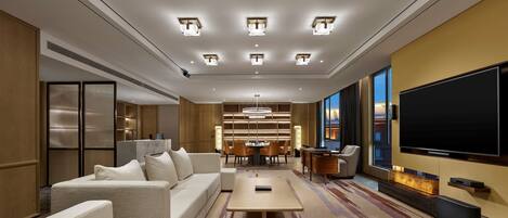 Quarto de hotel moderno com sofá, mesa de centro, televisão e área de refeições com cadeiras e mesa.