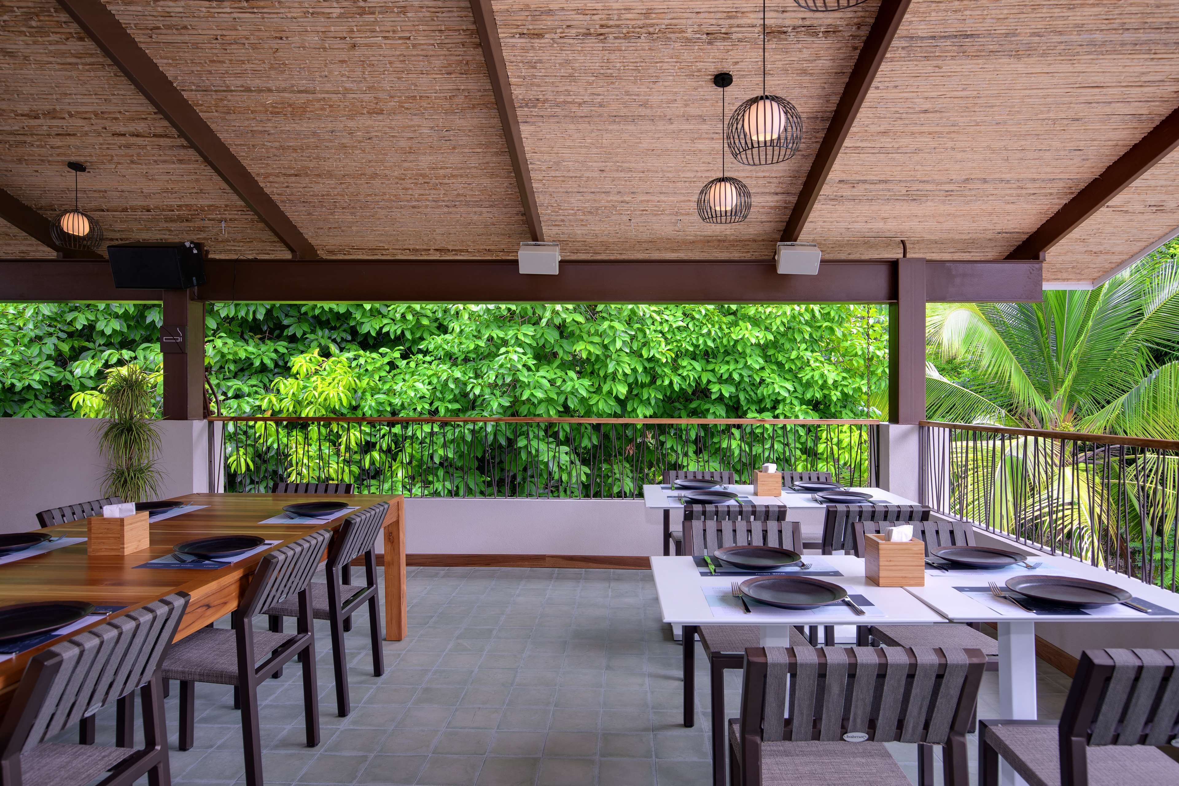 Um restaurante com mesas e cadeiras de madeira, piso de azulejos e vista para vegetação exuberante.