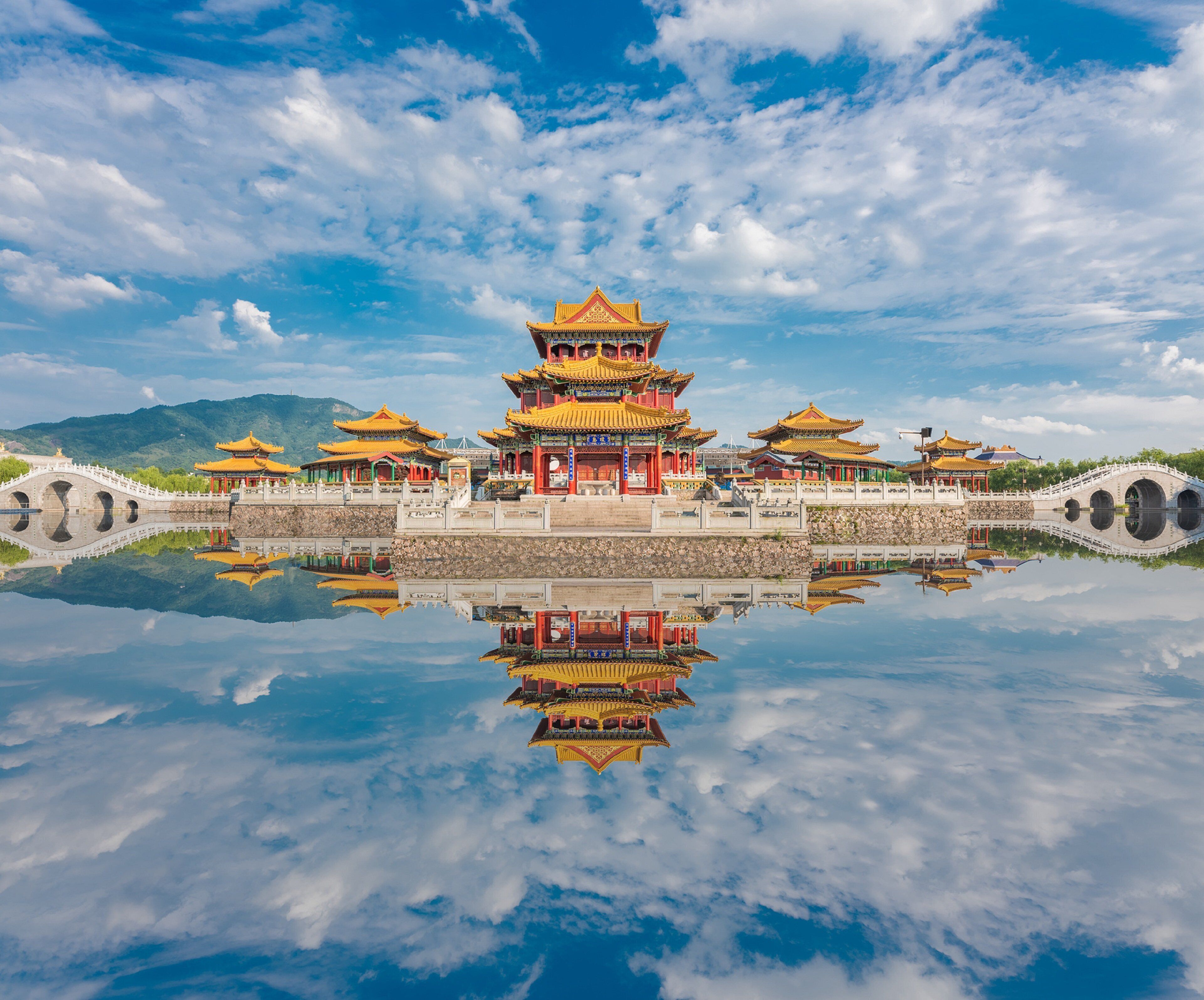 Um pavilhão chinês tradicional com vários níveis, refletindo em águas calmas, cercado por muros de pedra e uma ponte branca.