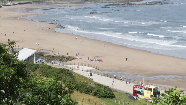 Uma praia costeira com areal, calçadão e um ônibus em primeiro plano.