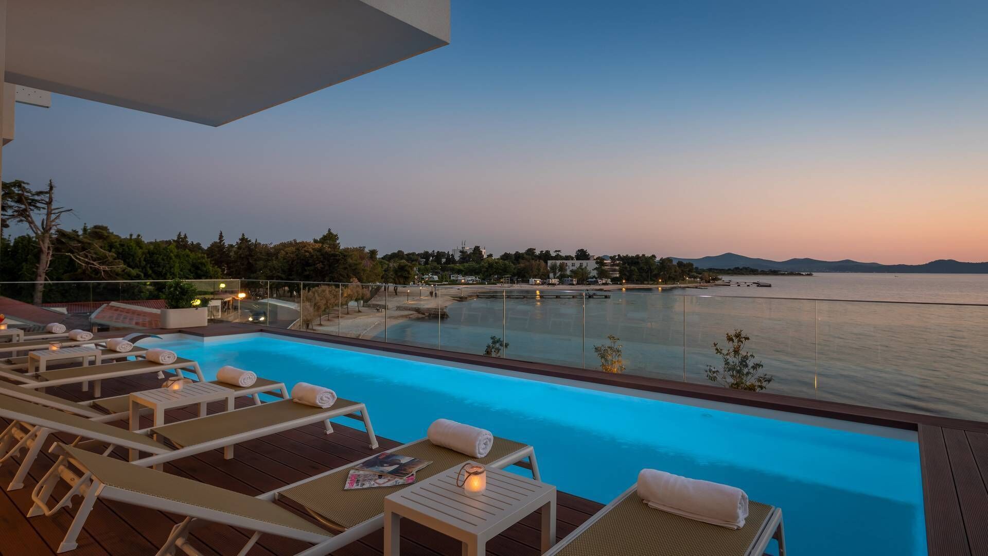 Área de piscina com espreguiçadeiras e uma mesa, com vista para um corpo de água e colinas distantes.