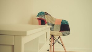 Uma cômoda branca com gavetas e um banquinho pequeno com almofada estampada.