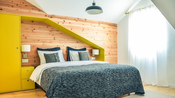 Quarto com teto inclinado de madeira, cama com colcha estampada escura, duas luminárias de cabeceira e um guarda-roupa amarelo.