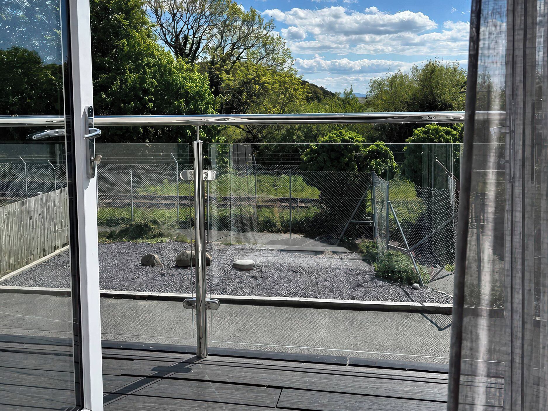 Uma estrutura de vidro com barras de metal, um deck de madeira e vista para uma área ajardinada com árvores e uma cerca.