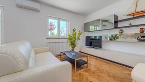 Uma sala de estar moderna com um sofá branco, piso de madeira em padrão herringbone, uma TV e um vaso com flores amarelas.
