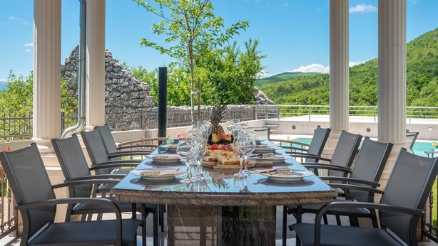 Uma área de refeições ao ar livre coberta, com uma mesa posta para refeição, cercada por cadeiras, com vista para uma paisagem deslumbrante com montanhas e uma piscina.