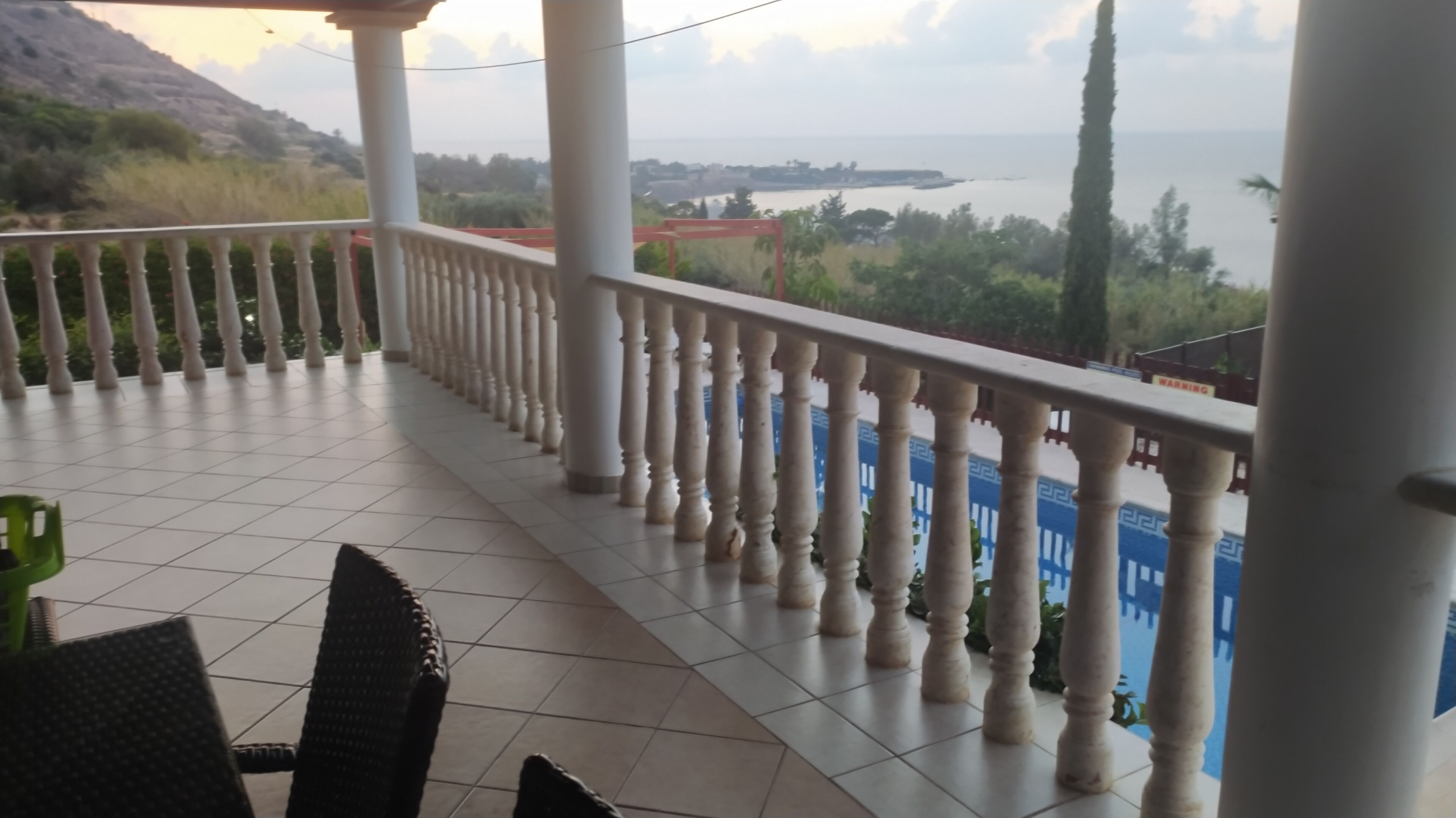 Uma varanda com balaústres brancos, soalho em azulejos e vista para um corpo de água e paisagem distante.