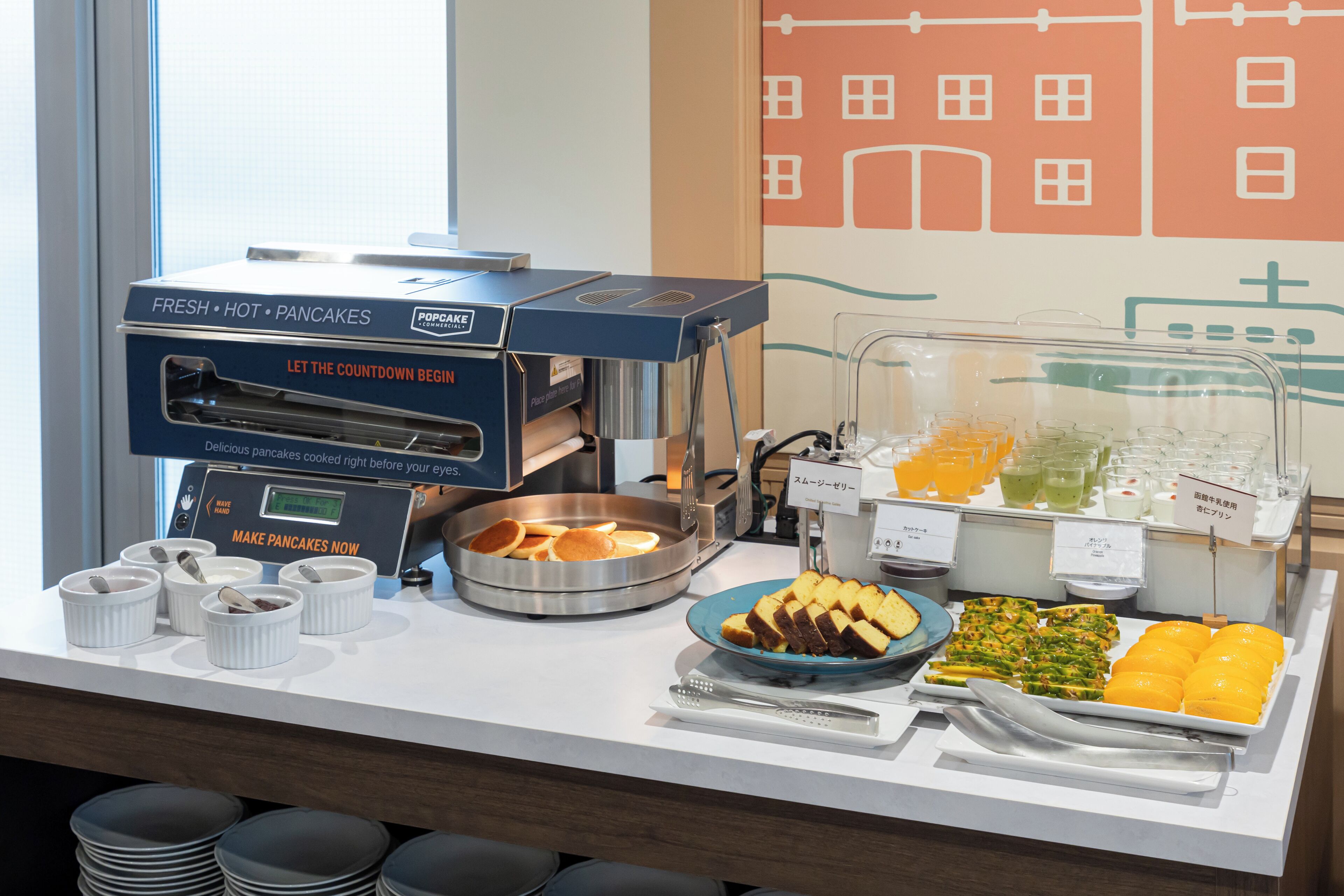 Buffet de café da manhã com máquina de panquecas, diversos pratos e dispensador de sucos.
