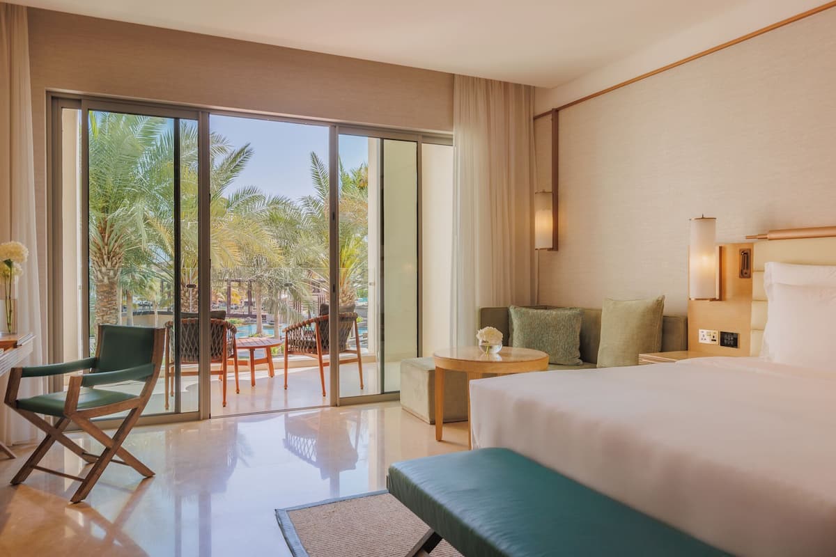 Quarto de hotel com uma cama grande, um sofá, uma mesinha e uma cadeira. Há uma porta de vidro deslizante que dá para uma área externa com piscina e palmeiras.