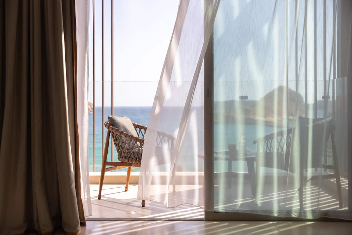 Quarto com vista para o mar, uma cadeira de vime e cortinas leves.