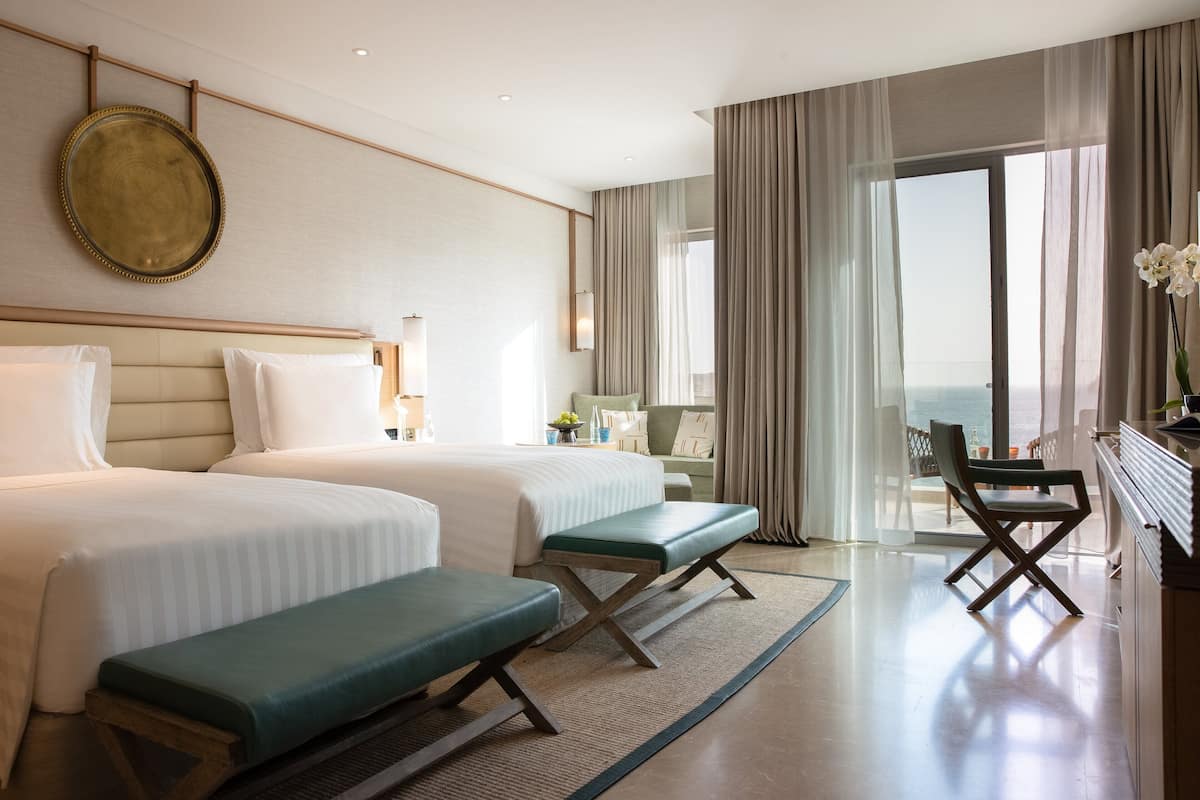 Quarto de hotel com duas camas, um banco, uma escrivaninha e vista para o oceano.