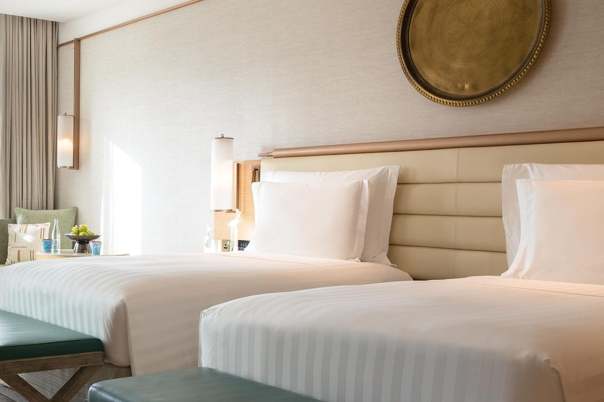 Quarto de hotel com duas camas, uma mesinha de cabeceira, um abajur e um prato decorativo na parede.