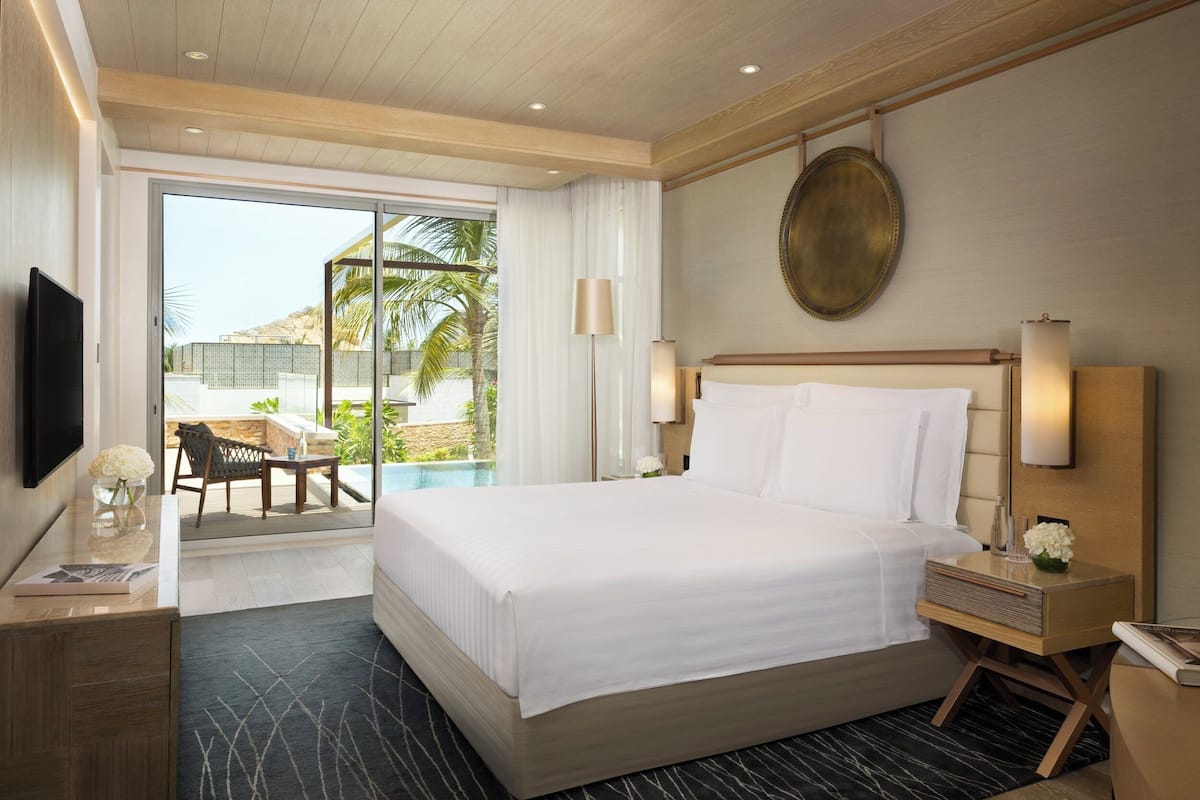 Quarto de hotel com uma cama grande, televisão, escrivaninha com cadeira e vista para uma piscina e palmeiras.