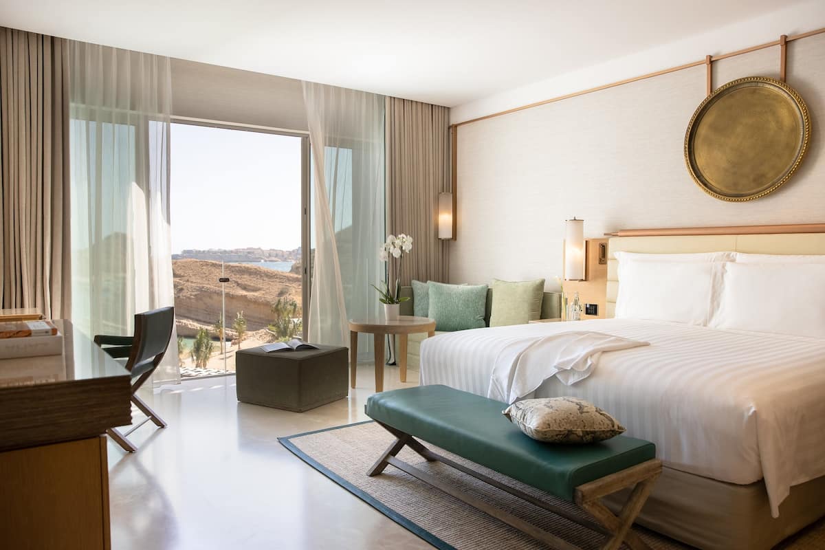 Quarto de hotel moderno com uma cama grande, área de estar com um banco, uma mesa pequena e vista para a paisagem.
