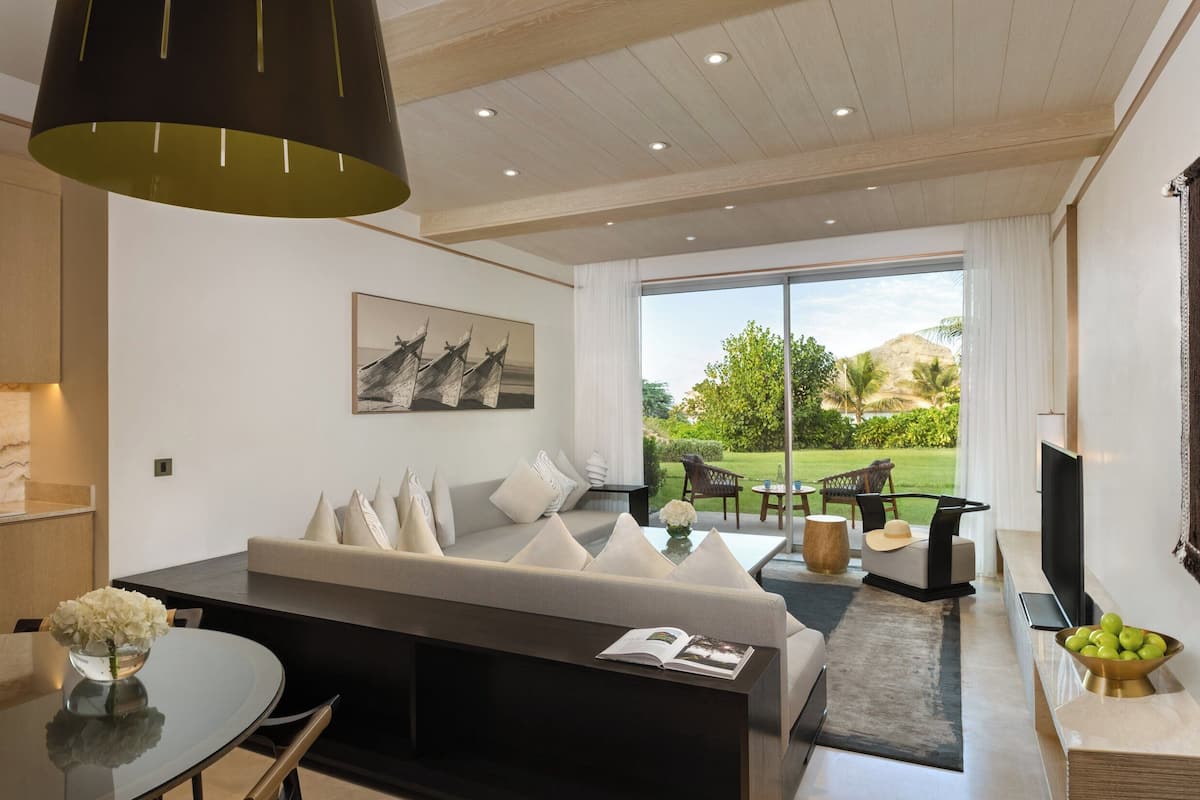 Uma sala de estar moderna com sofá, mesa de jantar e televisão. Há uma janela grande com vista para vegetação e colinas.