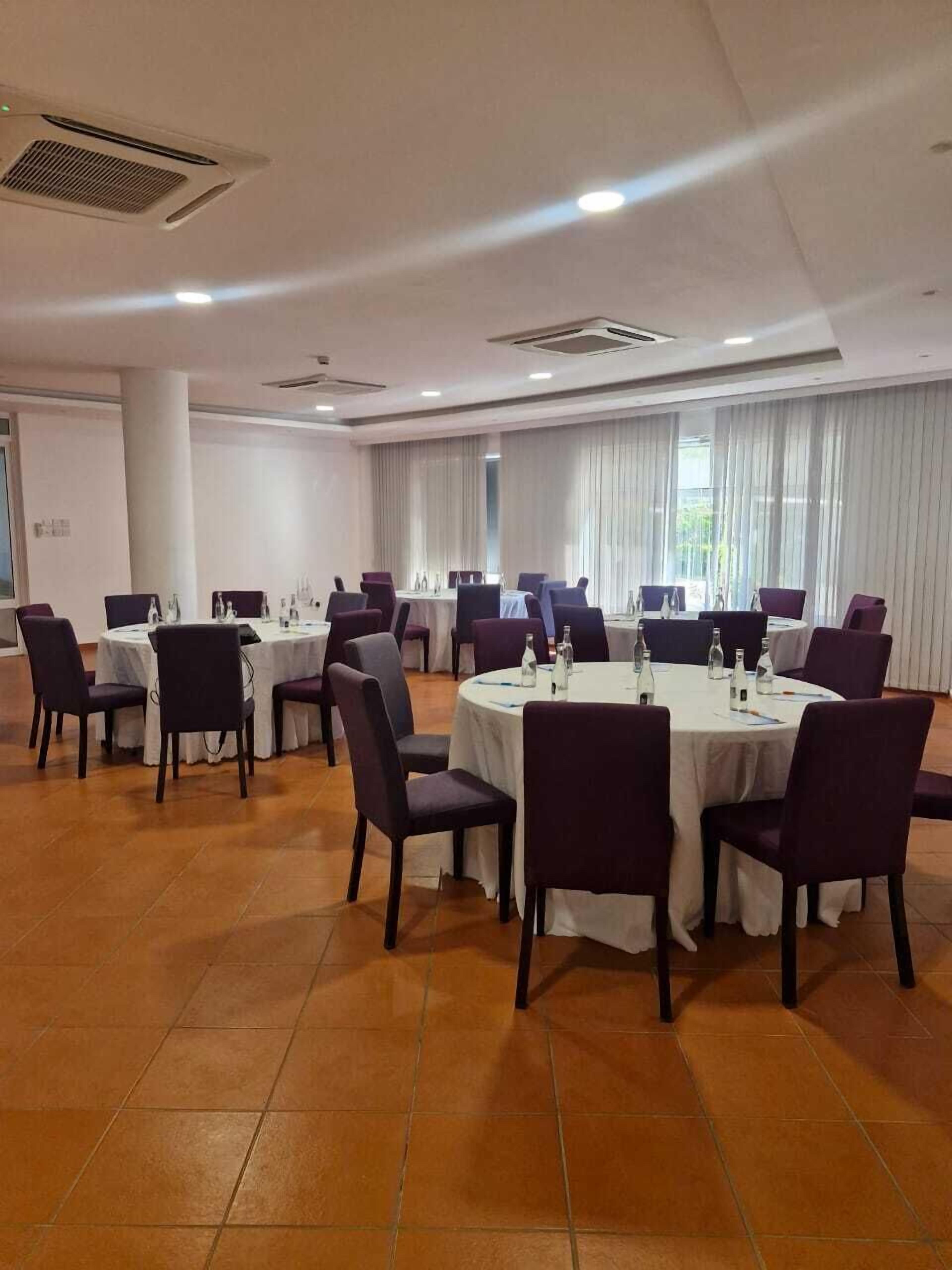 Uma sala de conferências com várias mesas redondas, cada uma rodeada por cadeiras. A sala possui grandes janelas com cortinas, iluminação embutida e piso revestido de azulejos.