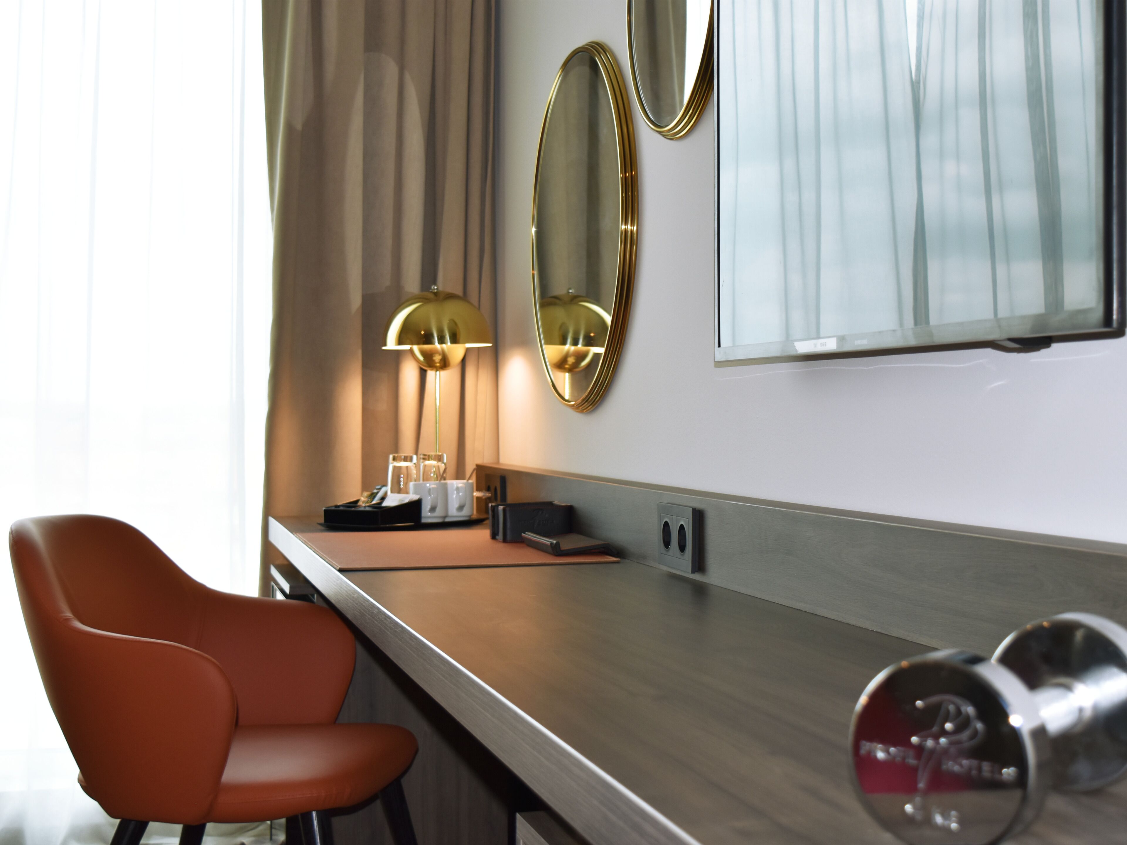 Quarto de hotel moderno com uma escrivaninha de madeira, uma cadeira vermelha, um abajur e um espelho.