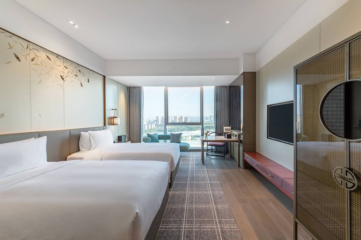 Quarto de hotel com duas camas, uma escrivaninha, uma televisão e vista para a cidade.