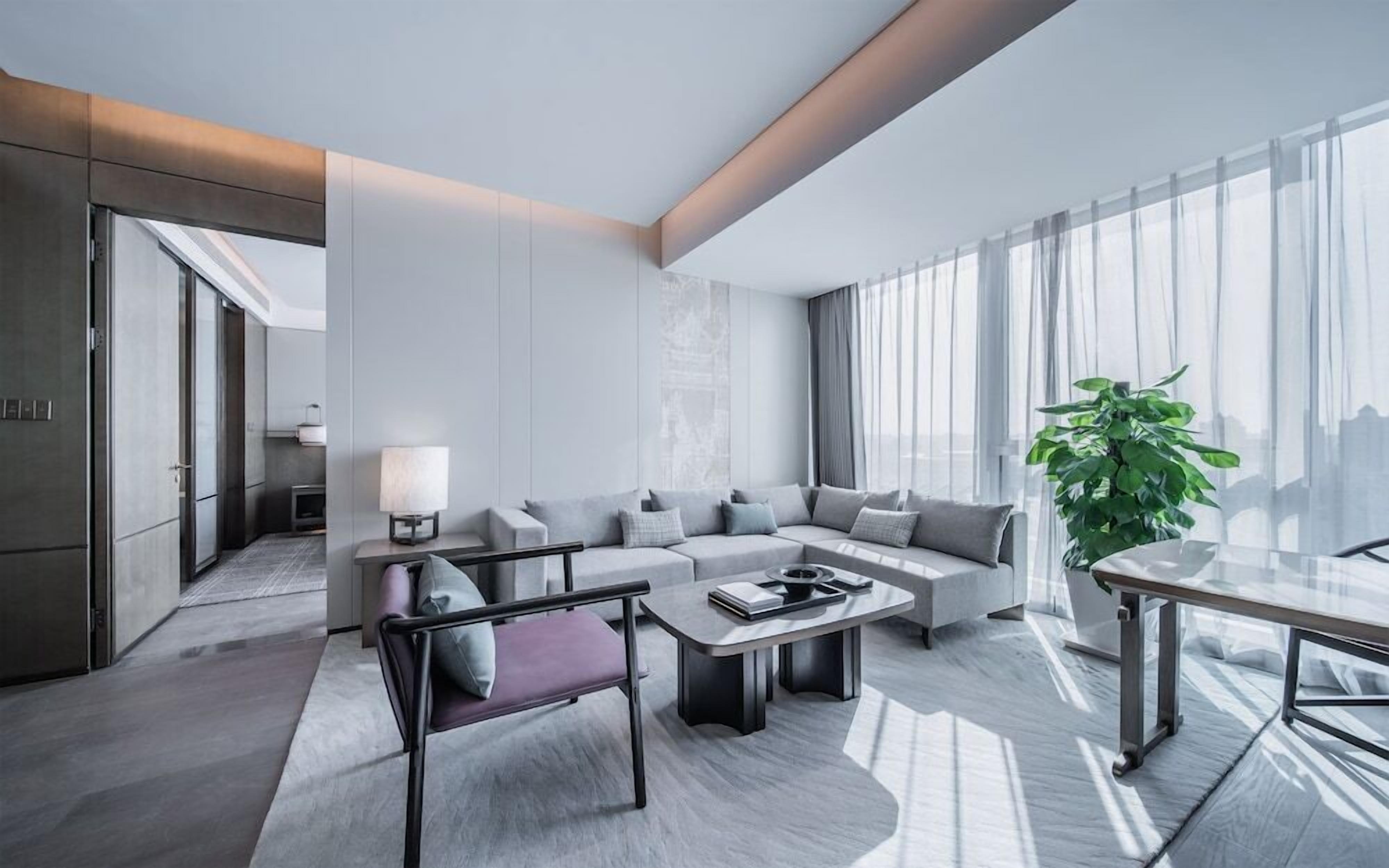Uma sala de estar moderna com um sofá modular cinza, uma mesa de centro e uma mesa de jantar. Há uma janela grande com cortinas leves e uma planta em vaso à direita.