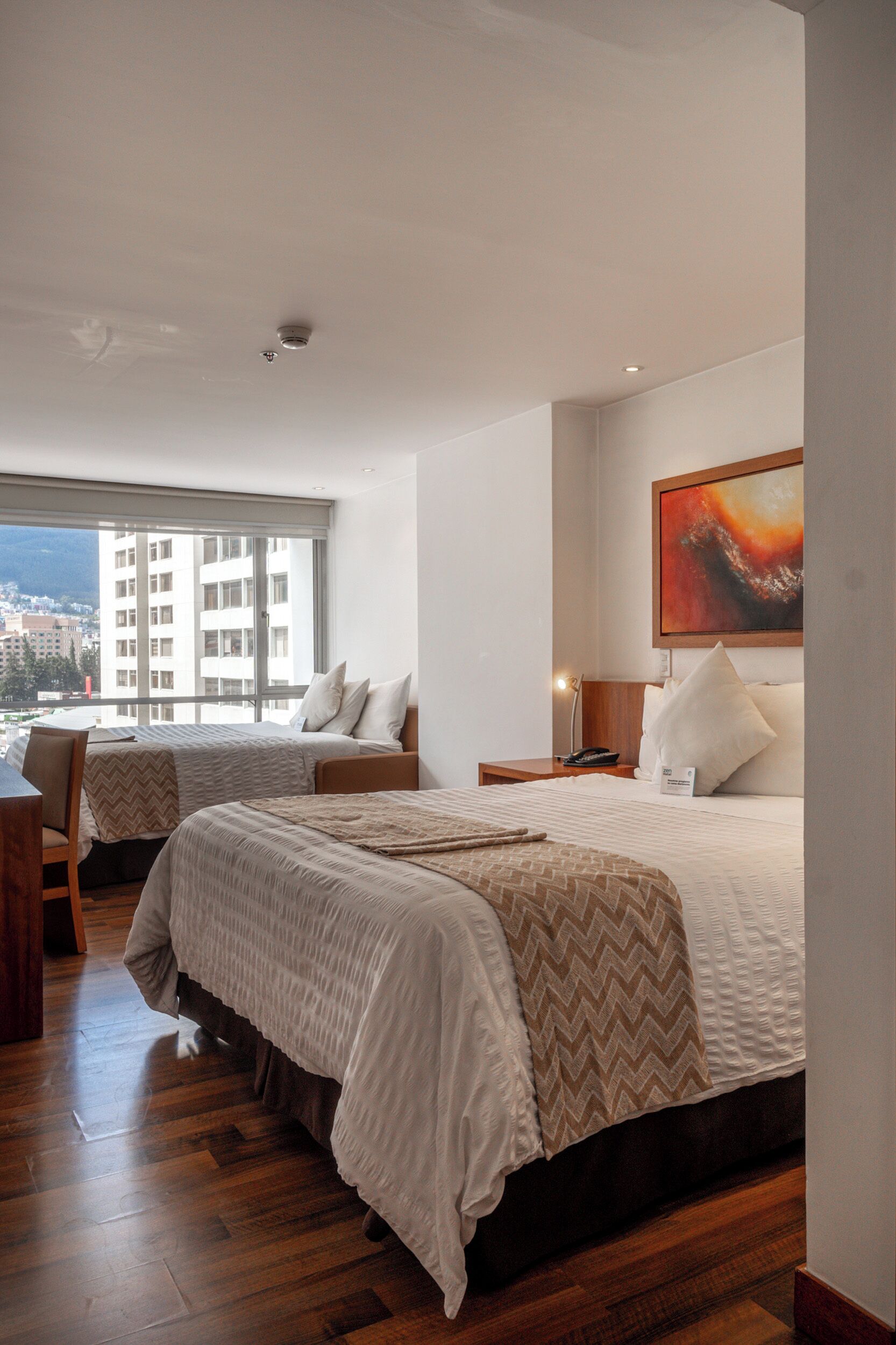Quarto de hotel moderno com duas camas, uma escrivaninha e uma janela grande com vista para a cidade.