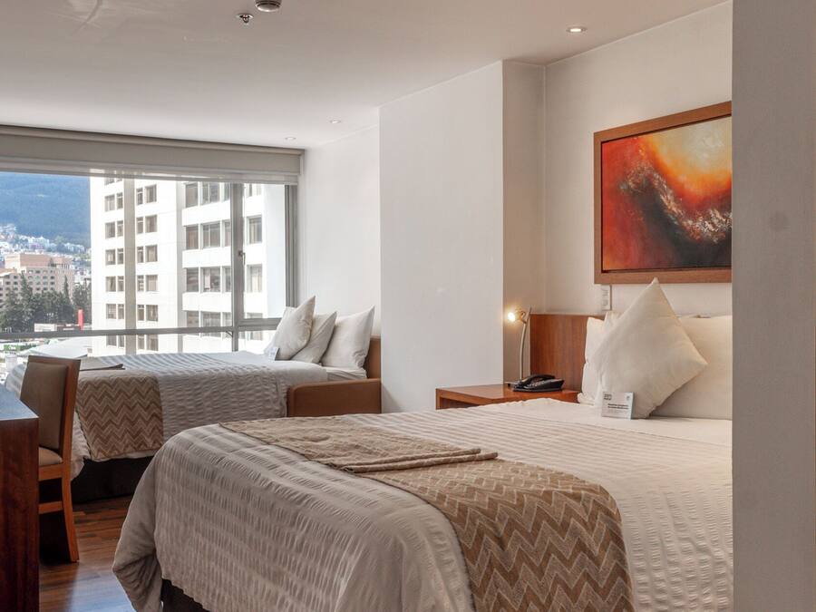 Quarto de hotel moderno com duas camas, uma escrivaninha e uma janela grande com vista para a cidade.